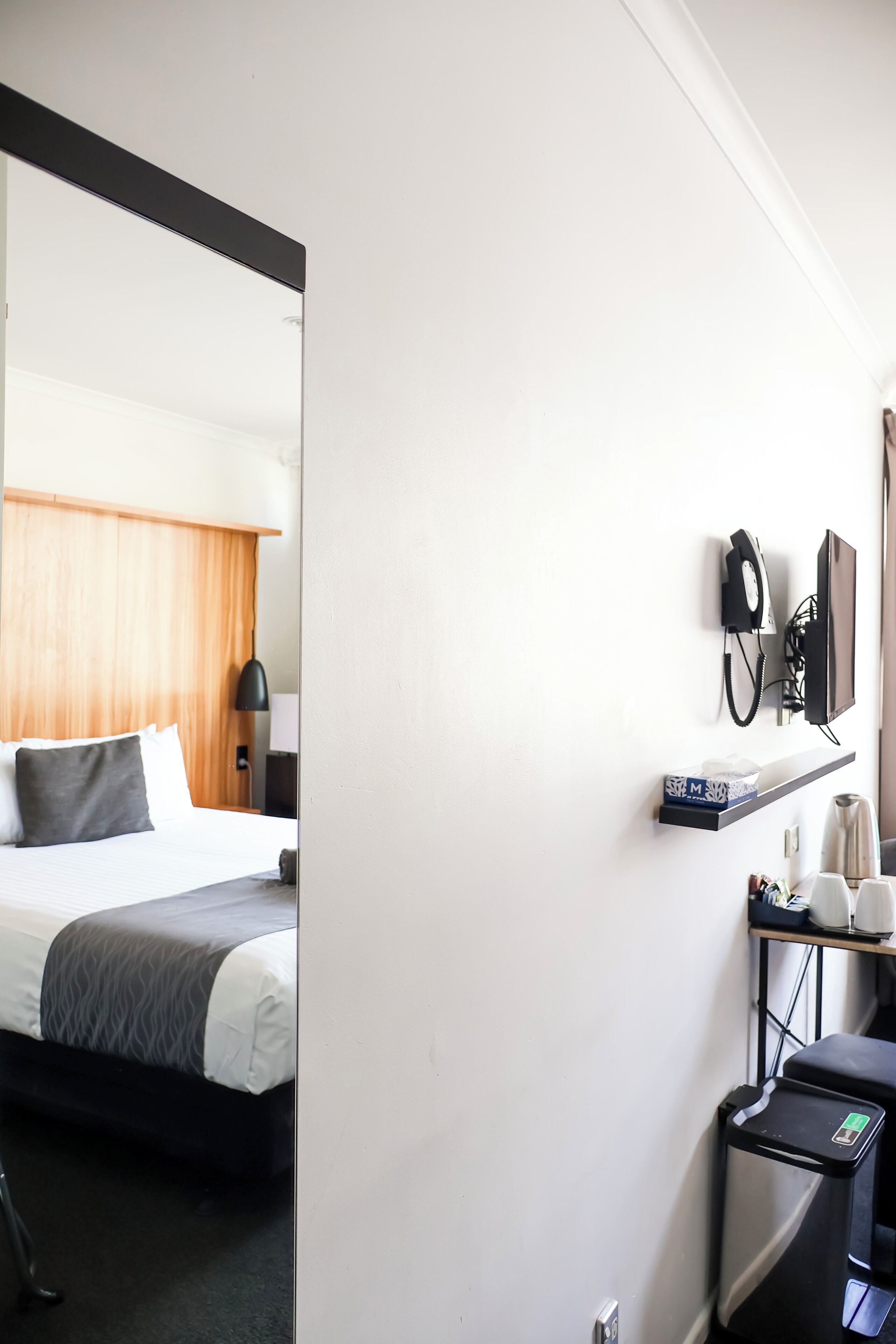 Quarto de hotel com cama, mesa de cabeceira com telefone e luminária, prateleira pequena com produtos de higiene pessoal e dispensador de água.