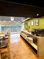 Interior moderno de café com mesas e cadeiras de madeira, um balcão com máquina de café e vista para o verde lá fora.