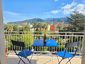 Varanda com duas cadeiras azuis dobráveis e uma mesa, com vista para uma paisagem urbana com montanhas e palmeiras.