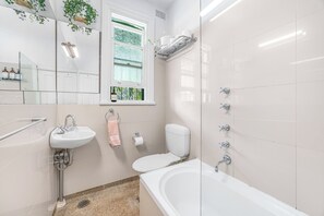 Uma casa de banho moderna com lavatório, sanita e banheira brancos. Há uma janela com vista para a vegetação lá fora e uma prateleira com artigos de higiene pessoal.