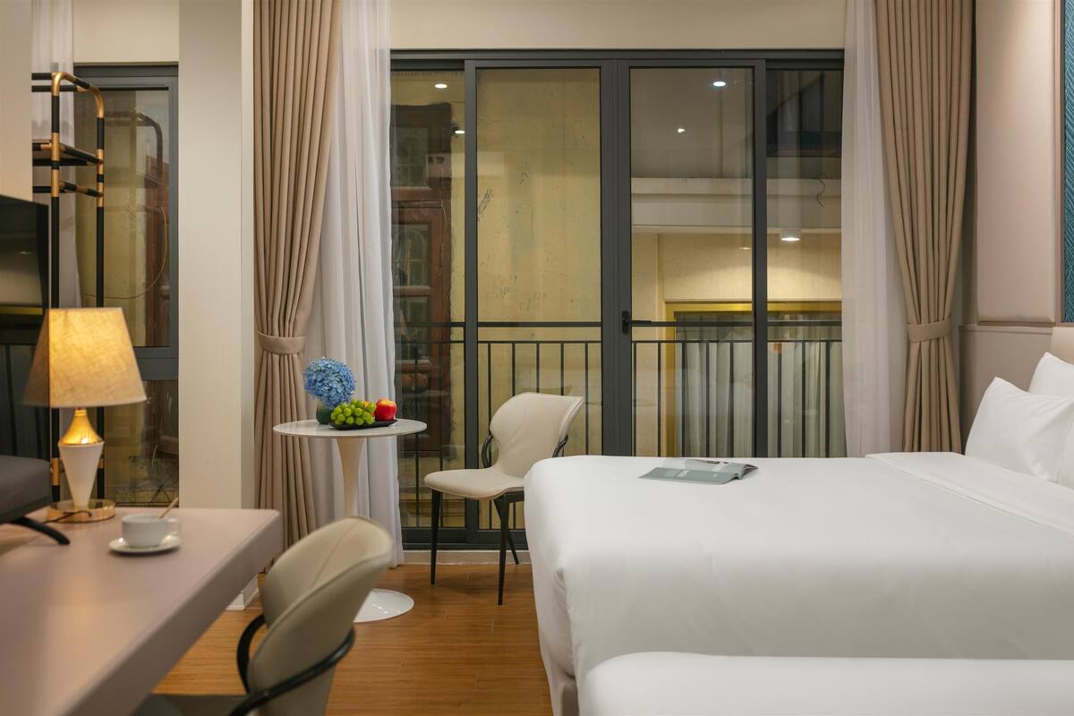 Quarto de hotel com cama, escrivaninha com luminária, cadeira, mesinha com frutas e varanda com cortinas.
