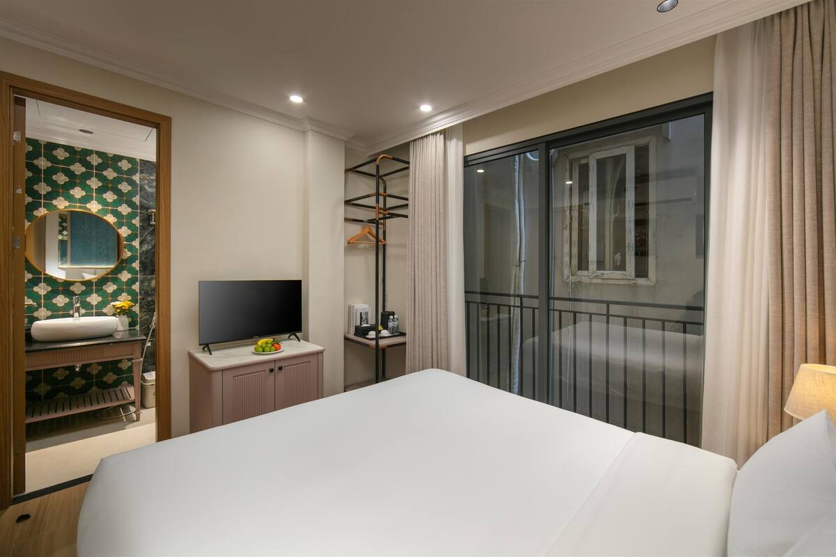 Quarto de hotel com cama, televisão, banheiro com espelho e varanda com cortinas.