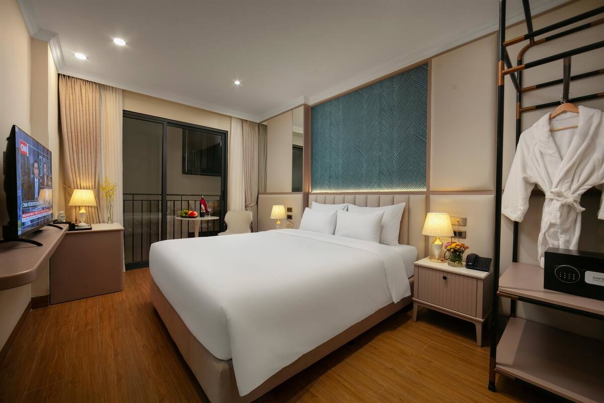 Quarto de hotel com cama grande, mesas de cabeceira com abajures, televisão, guarda-roupa e varanda com vista.