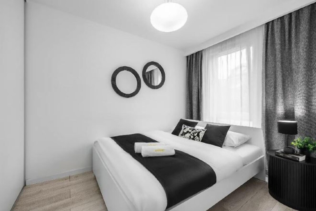 Quarto moderno com cama, dois espelhos fixados na parede, mesa de cabeceira com abajur e janela com cortinas.
