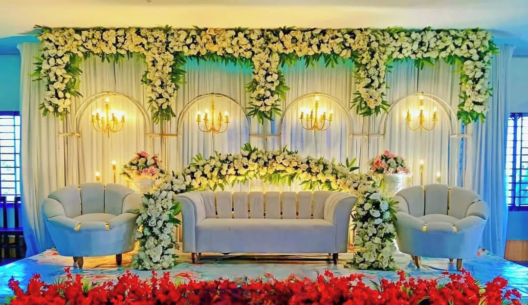 Um palco com um sofá e duas poltronas, cercado por arranjos florais e cortinas.