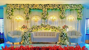 Um palco com um sofá e duas poltronas, cercado por arranjos florais e cortinas.