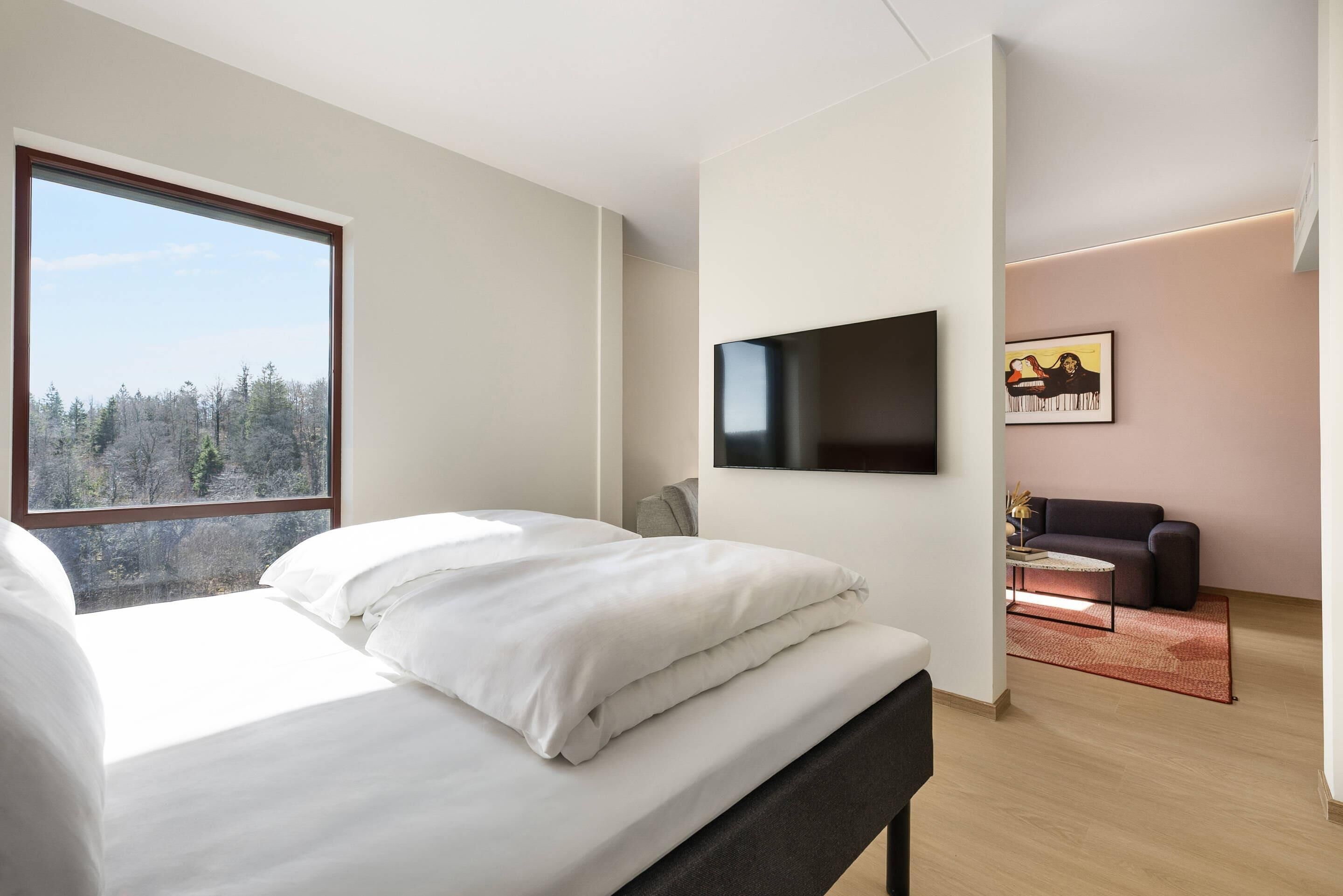 Quarto de hotel moderno com uma cama grande, televisão na parede e vista para a floresta pela janela.