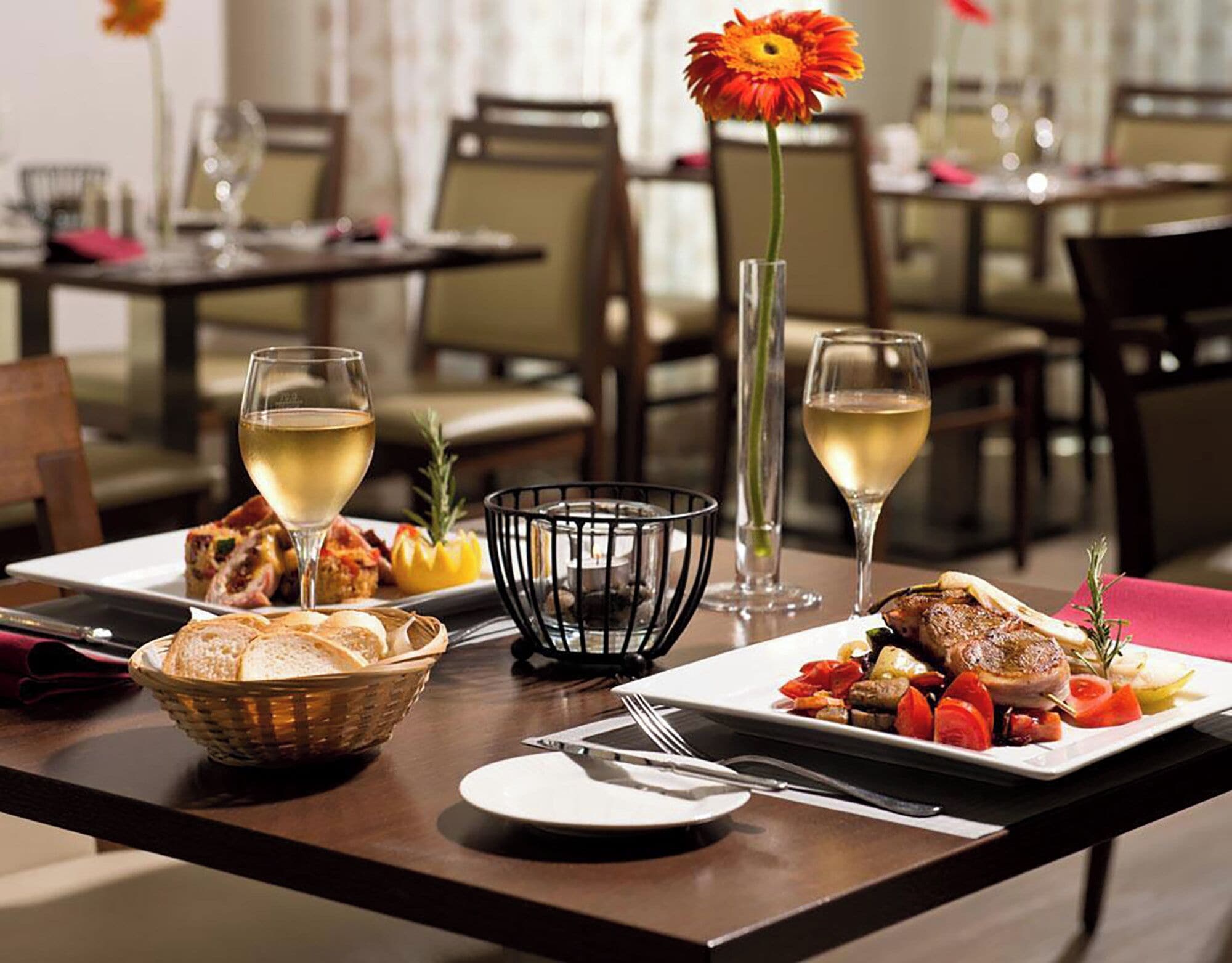 Uma mesa de jantar posta com uma cesta de pão, um prato de carne assada com legumes, duas taças de vinho branco e um vaso com uma única flor de laranja.