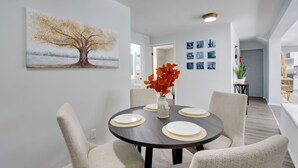 Uma área de refeições com uma mesa redonda, cadeiras brancas e um vaso com flores vermelhas. Há uma pintura de uma árvore na parede e várias fotos emolduradas na parede ao lado.