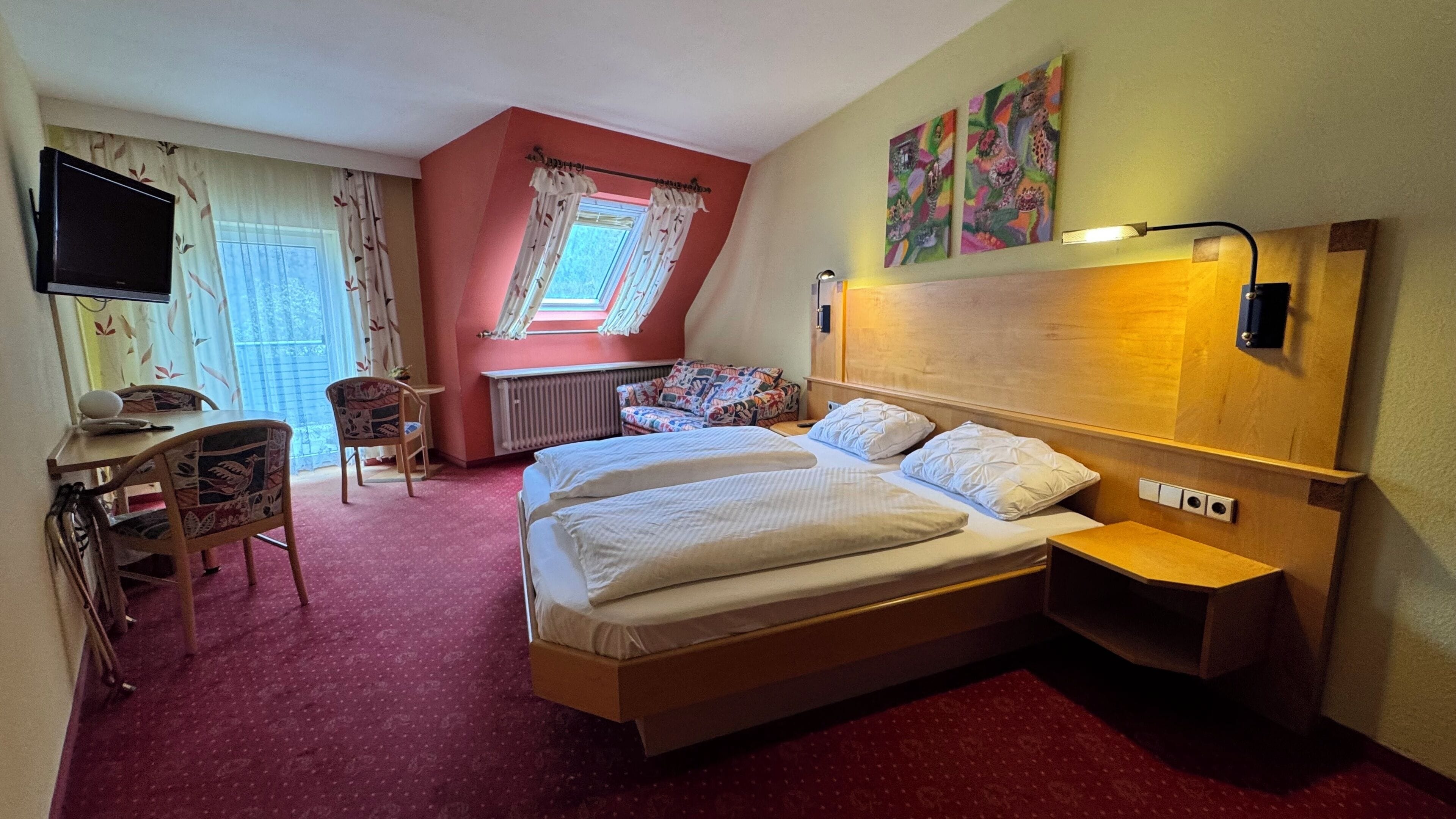 Quarto de hotel com uma cama grande, uma escrivaninha, uma cadeira, uma televisão e uma janela com cortinas.