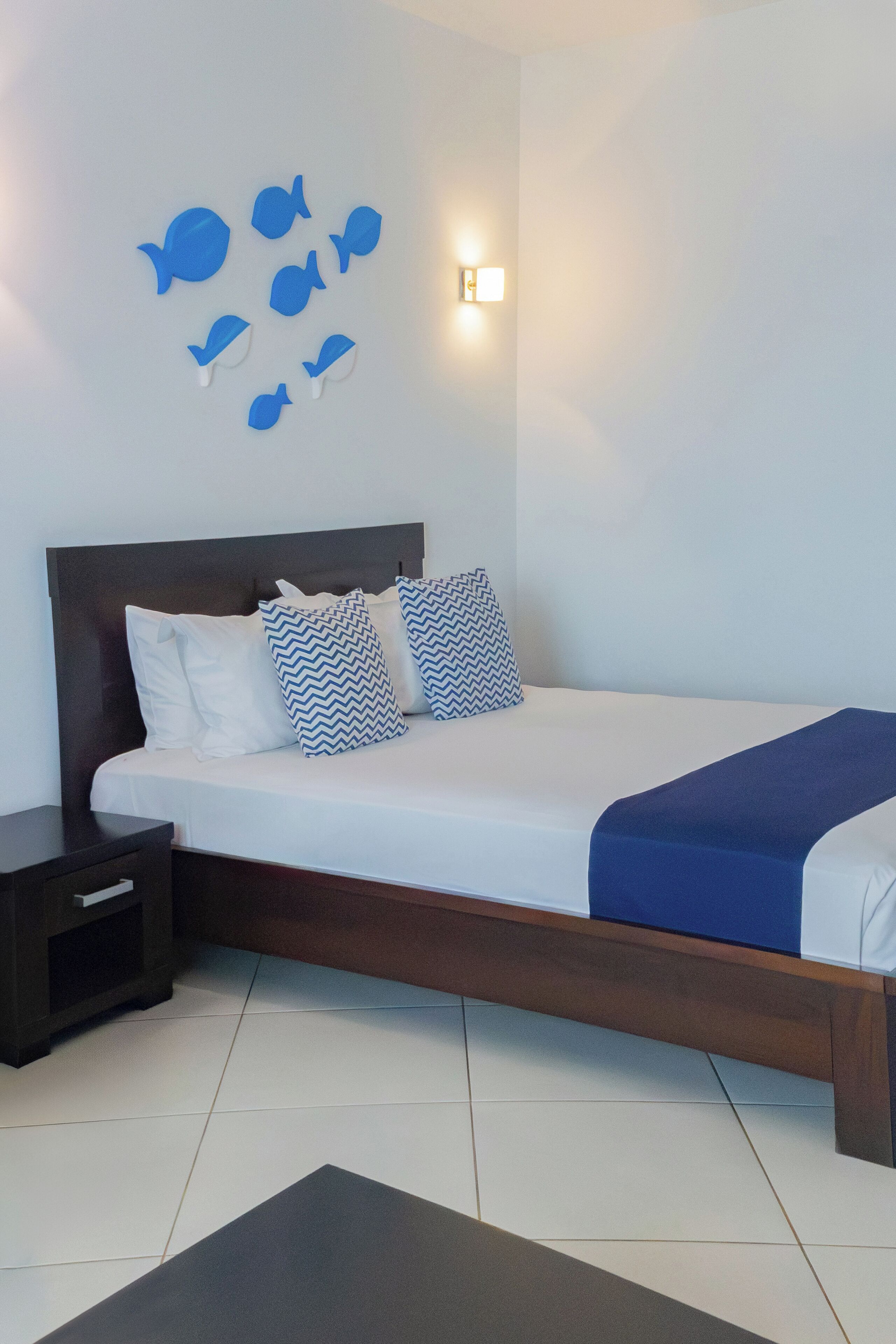 Uma cama bem arrumada com almofada estampada em azul e branco, cabeceira de madeira escura e luminária fixada na parede.