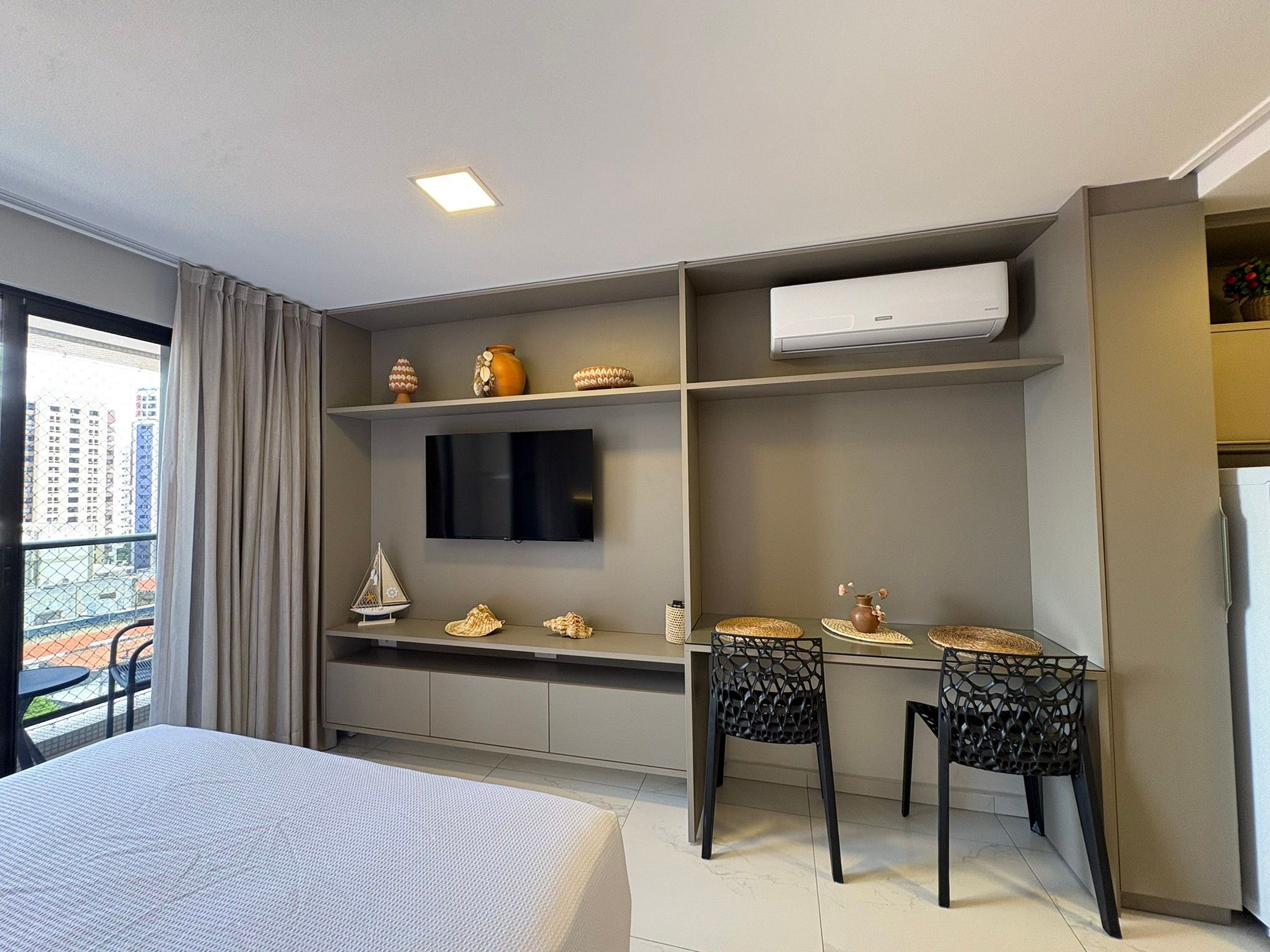 Quarto de hotel com cama, escrivaninha com duas cadeiras, televisão de tela plana e ar condicionado integrado.