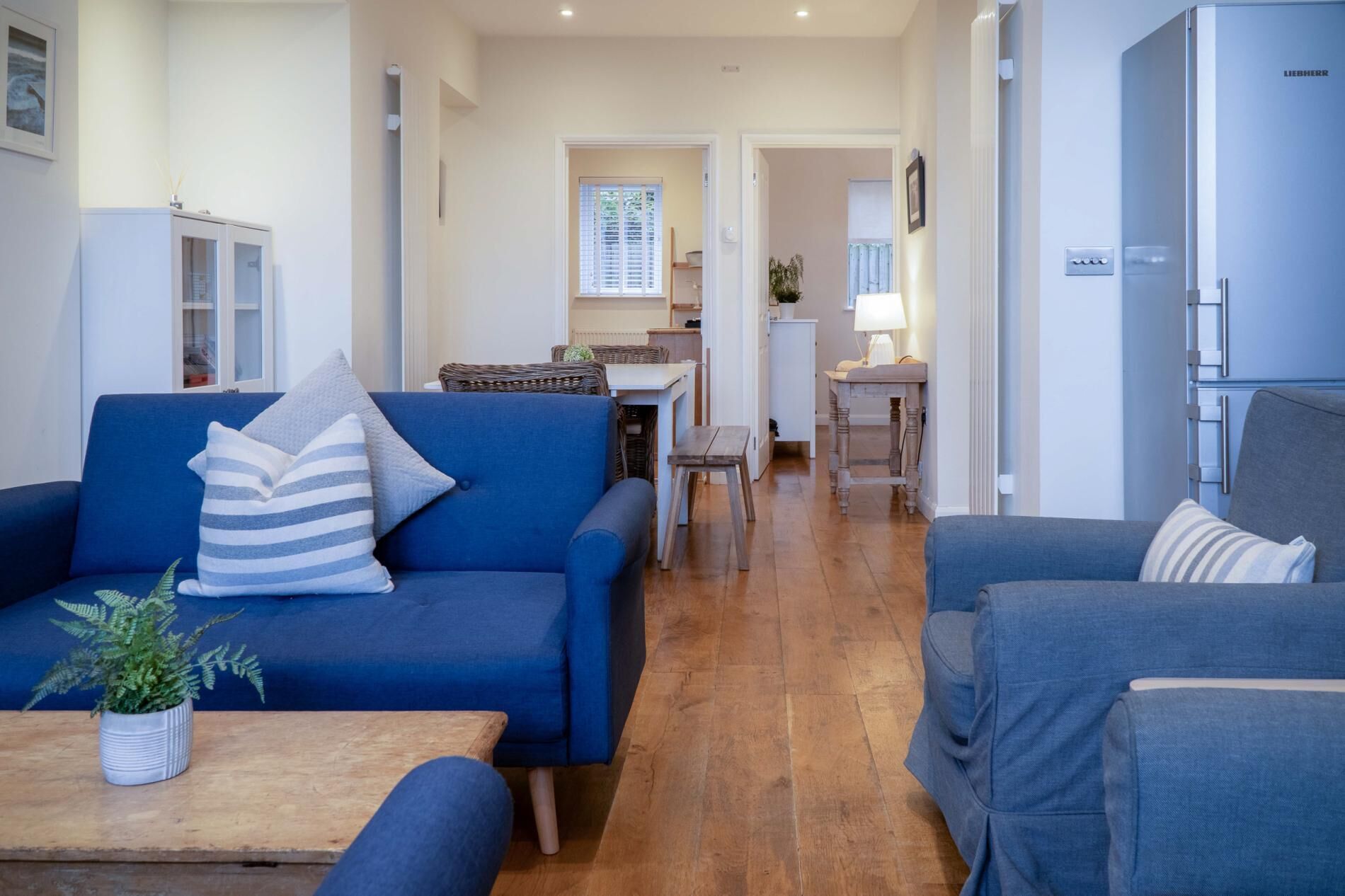 Uma sala ampla com um sofá azul, uma poltrona menor e uma mesa de centro de madeira. Uma mesa de jantar com cadeiras é visível ao fundo.