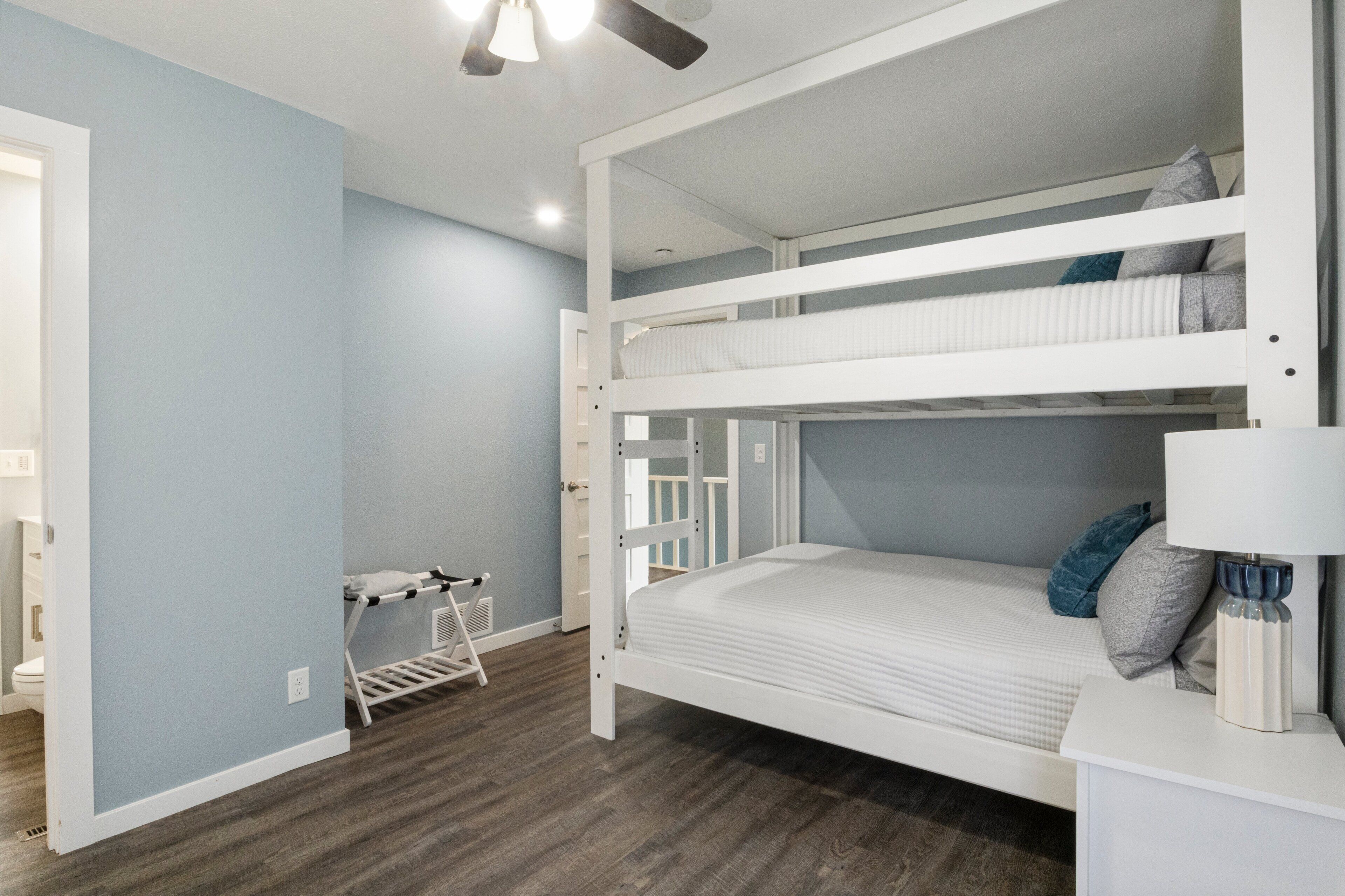 Quarto com beliche, cabeceira branca, mesinha de cabeceira com abajur e piso de madeira.