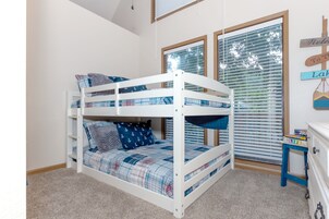Uma cama beliche com roupa de cama xadrez, uma mesinha com um livro e uma janela com persianas.