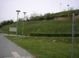 Uma passarela pavimentada ao lado de uma colina gramada, com postes de iluminação e uma placa de faixa de pedestres.