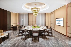 Sala de conferências com mesa redonda preparada para uma reunião, um candelabro e uma tela de projeção exibindo informações do hotel.