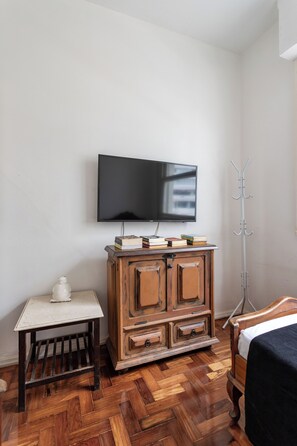 Quarto com um armário de madeira, televisão na parede, uma mesinha com um vaso e uma cama com cobertura escura.