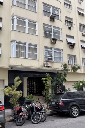 Um edifício de vários andares com fachada bege, diversas janelas com molduras brancas e aparelhos de ar condicionado. Duas motocicletas estão estacionadas na frente, e um SUV preto também é visível.