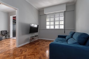 Uma sala de estar com um sofá azul, uma televisão de tela plana, piso de madeira em padrão herringbone e uma janela com persianas.