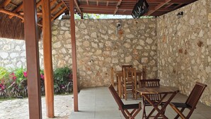 Área de varanda coberta com parede de pedra, móveis de madeira e luminárias penduradas.