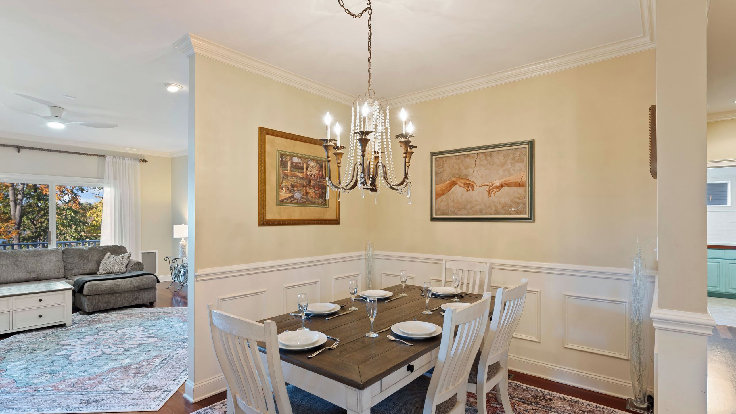 Uma sala de jantar com mesa de madeira posta para seis pessoas, cadeiras brancas, um candelabro e dois quadros na parede.