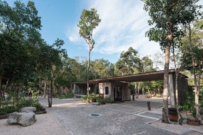 Um edifício rústico com varanda e acesso coberto, rodeado por árvores e caminhos de cascalho.