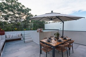 Terraço no telhado com mesa de jantar posta para seis pessoas, rodeado por cadeiras de vime, um guarda-sol grande e um sofá branco com almofadas.