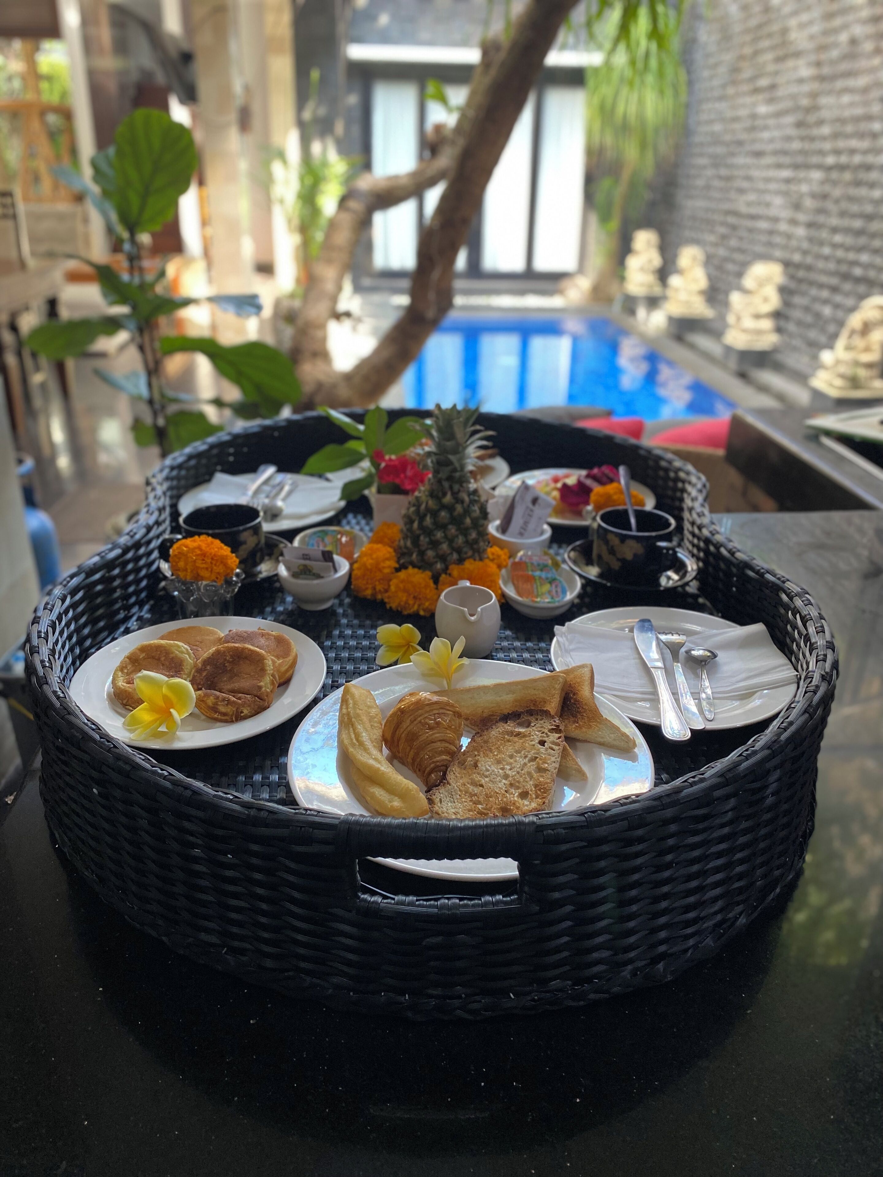Uma bandeja de vime com itens de café da manhã, incluindo croissants, pão e uma abacaxi, colocada sobre uma mesa com vista para um edifício e plantas ao fundo.