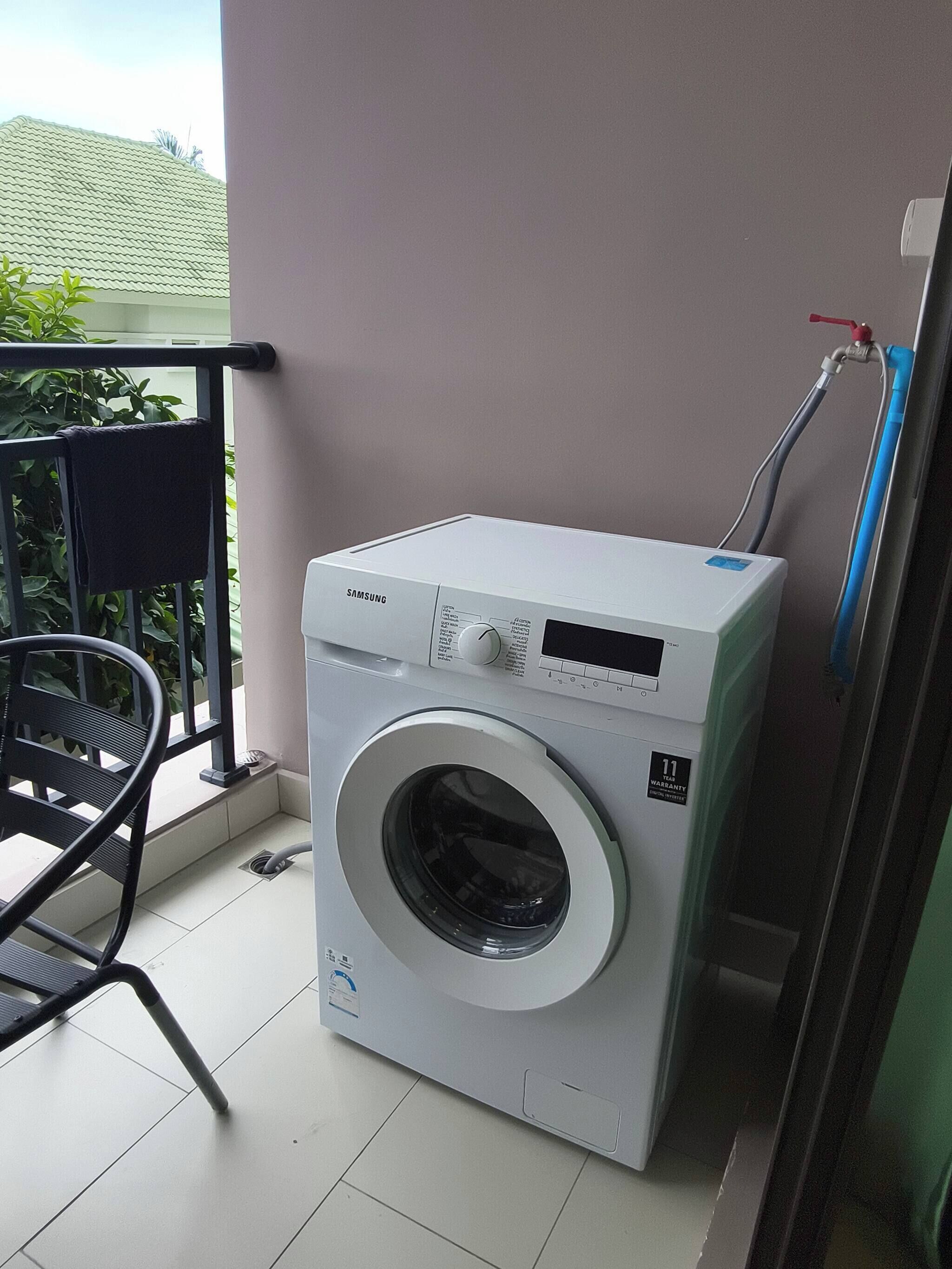 Uma máquina de lavar Samsung numa varanda, com uma cadeira e uma mangueira.