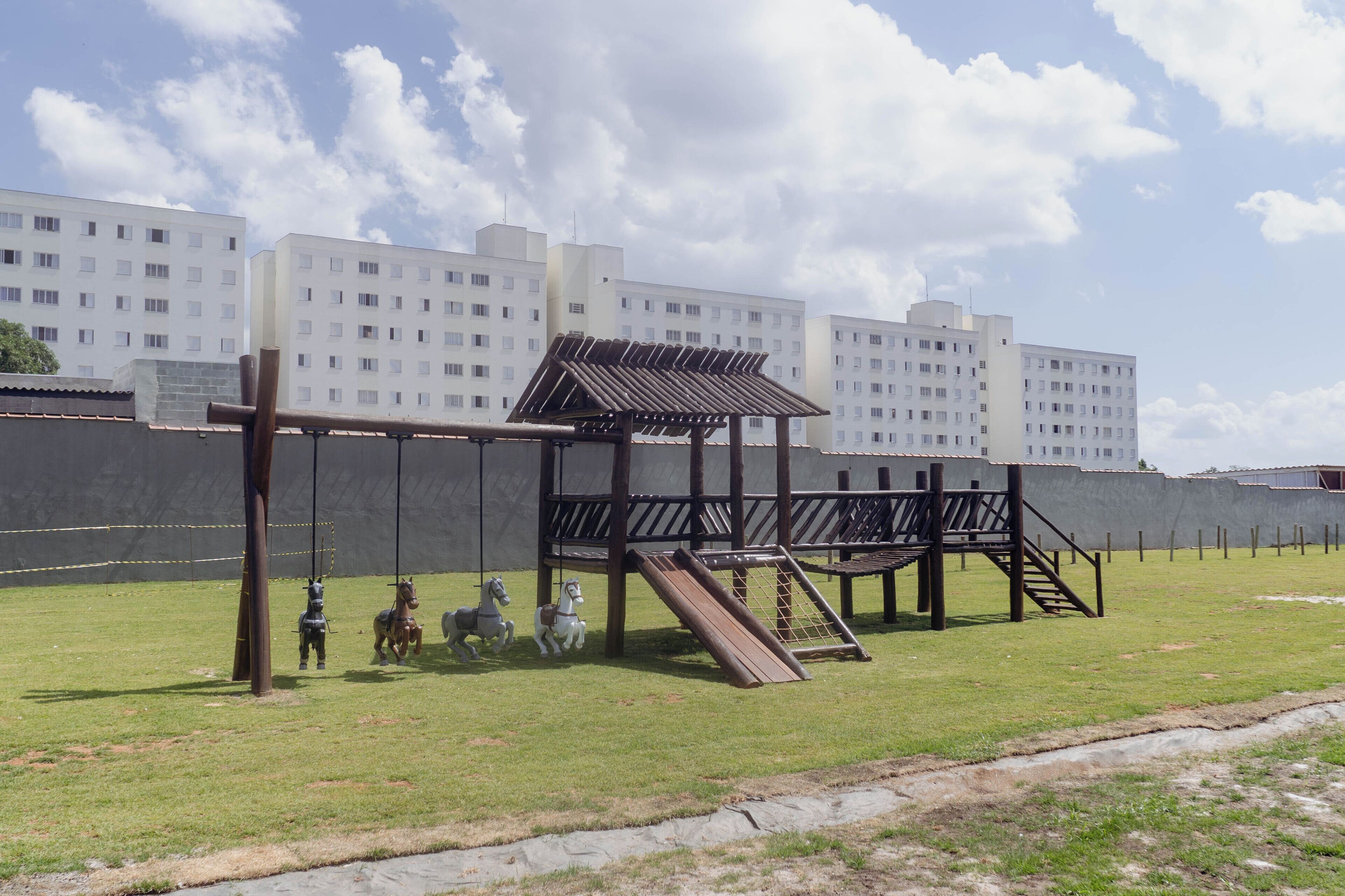 Um parquinho com estrutura de madeira, incluindo um escorrega e área de escalada, com prédios residenciais ao fundo.