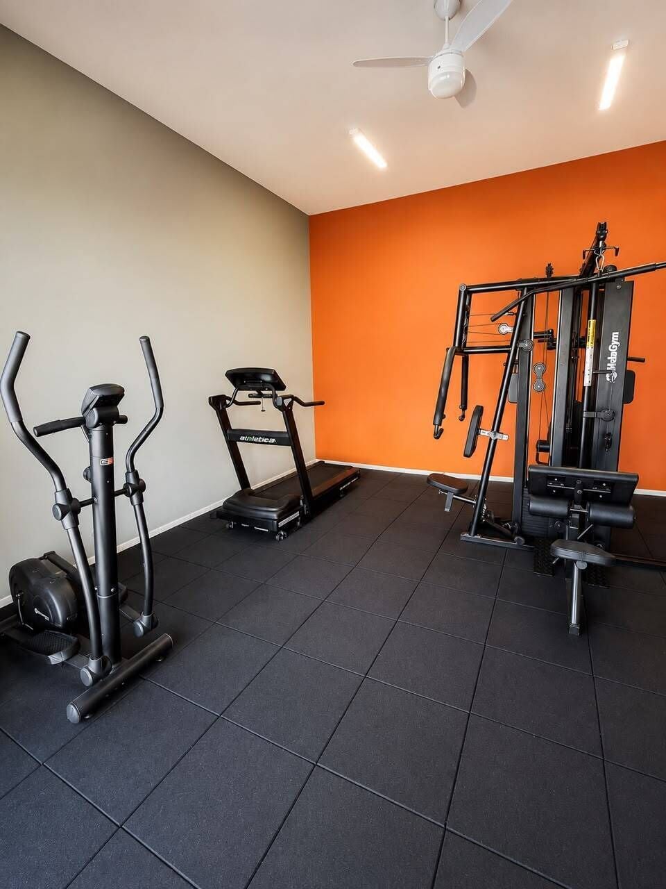 Academia com esteira, elíptica e aparelhos de musculação em frente a uma parede laranja.