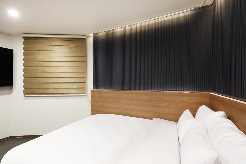 Quarto de hotel moderno com cabeceira de madeira, uma cama grande com roupa de cama branca, painéis de parede escuros e uma janela com persianas horizontais.