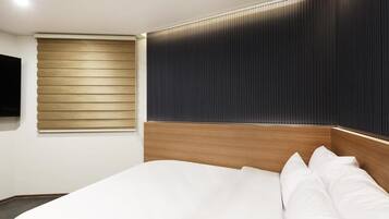 Quarto de hotel moderno com cabeceira de madeira, uma cama grande com roupa de cama branca, painéis de parede escuros e uma janela com persianas horizontais.