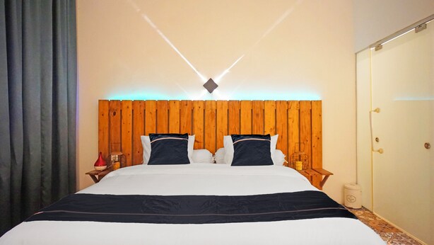 Quarto de hotel com cabeceira de madeira, cama com roupa de cama branca e escura, mesa de cabeceira com abajur e uma porta à direita.
