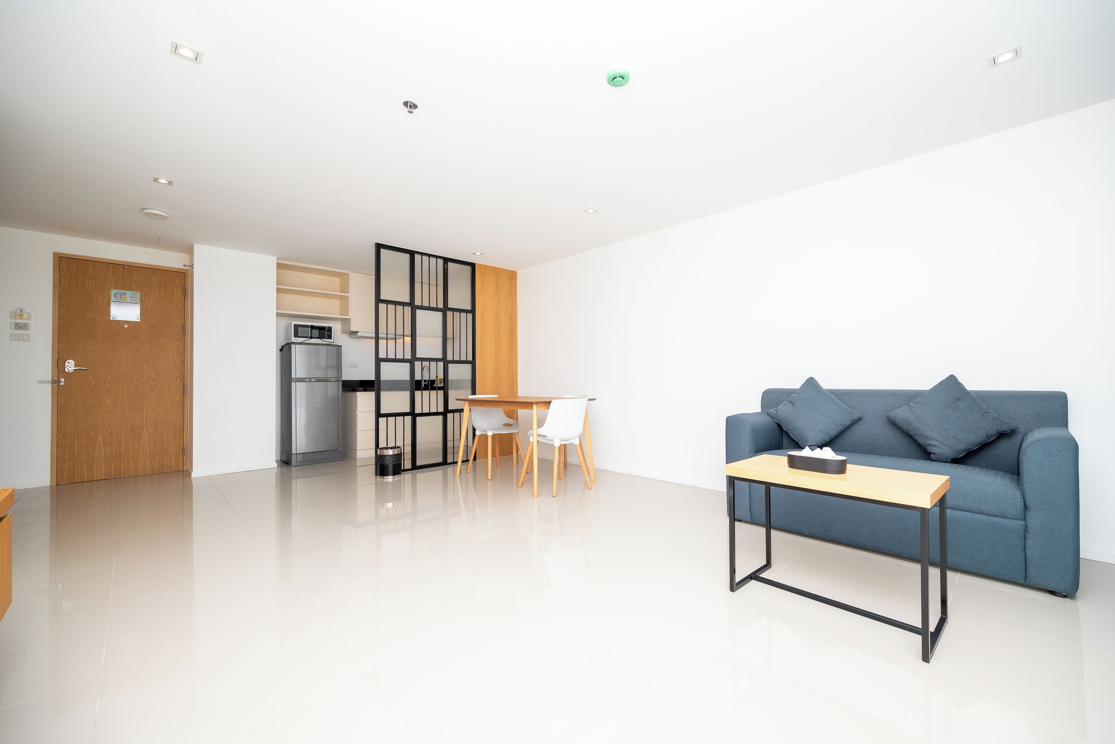 Uma sala de estar moderna com um sofá azul, uma mesa de centro de madeira e uma área de cozinha com geladeira e mesa de jantar.