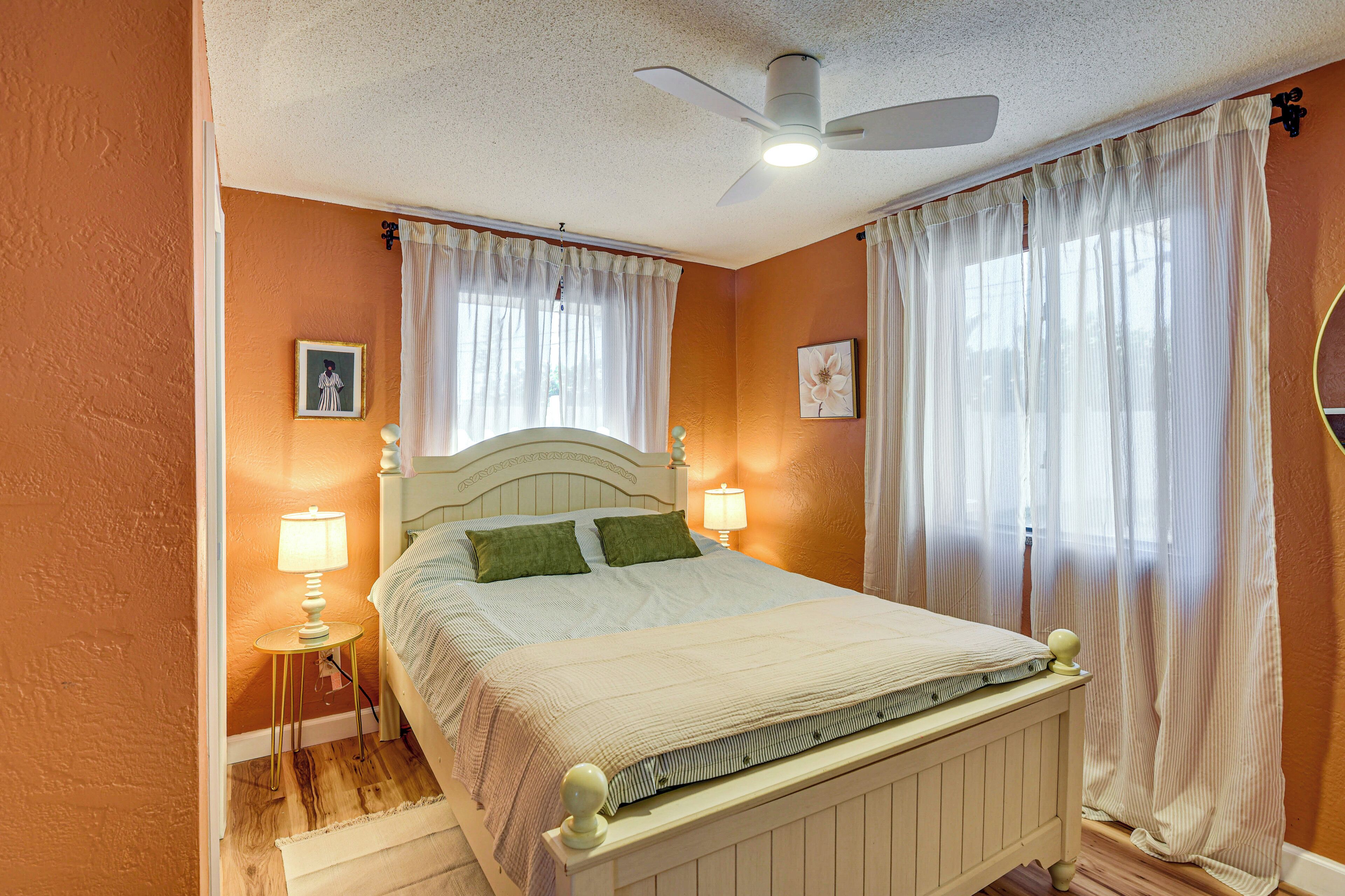 Quarto com cama, mesa de cabeceira, ventilador de teto e duas janelas com cortinas finas.