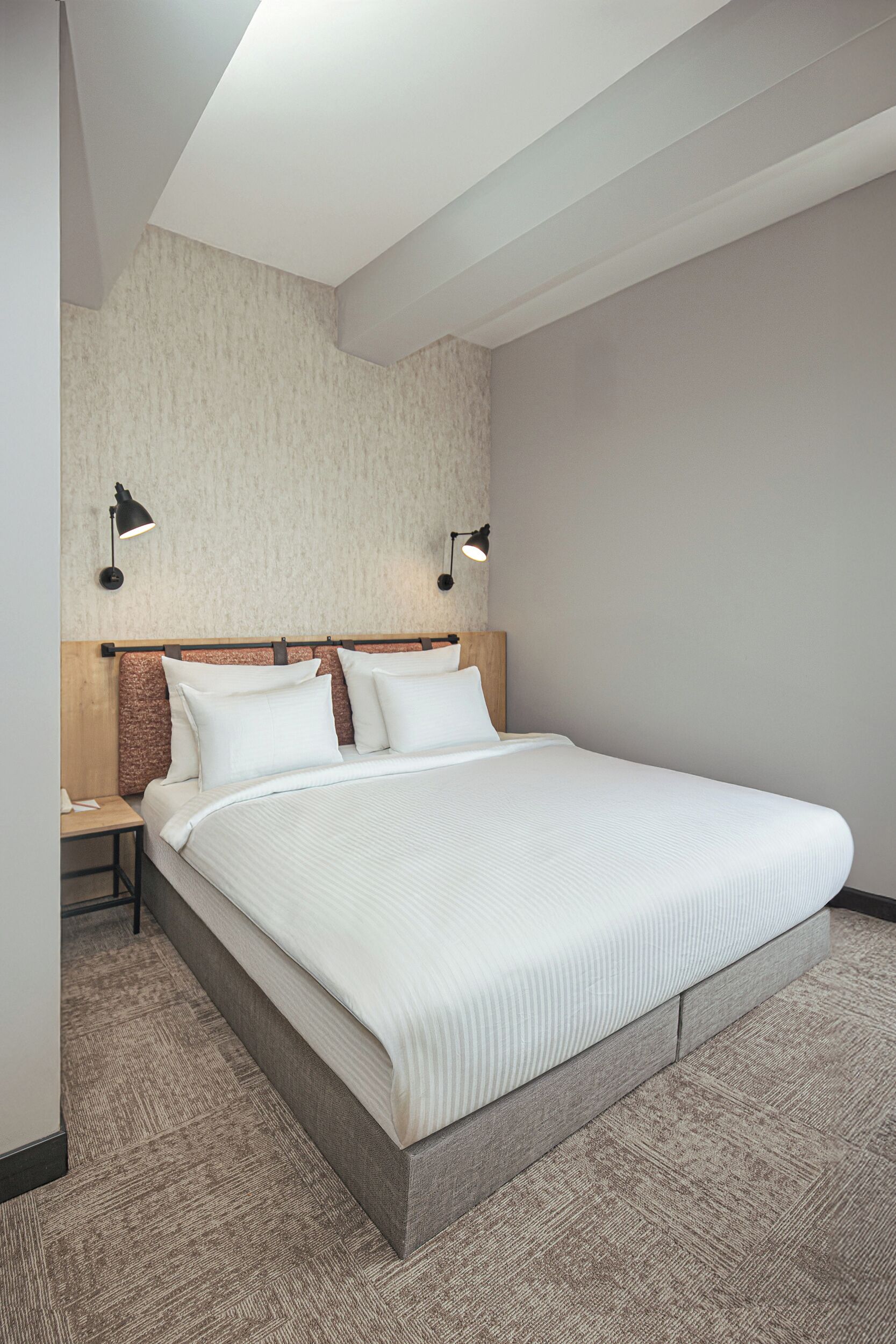 Quarto de hotel moderno com uma cama grande, duas mesinhas de cabeceira e luminárias fixadas na parede.
