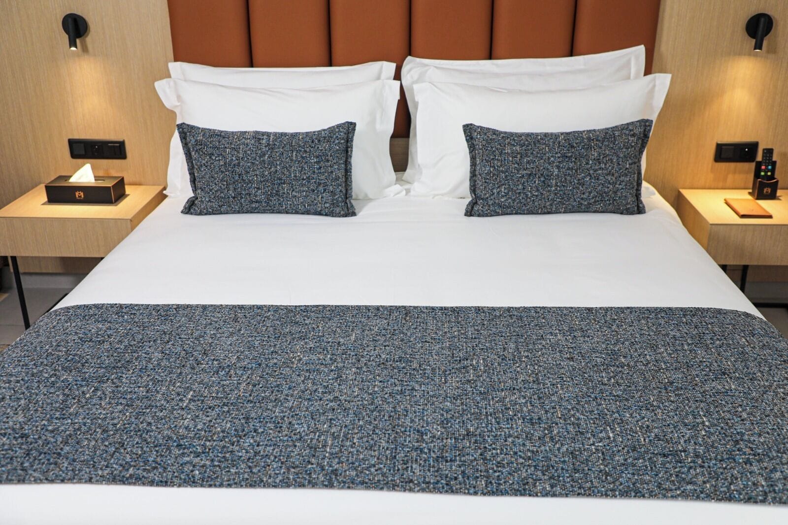 Quarto de hotel com uma cama grande, dois travesseiros brancos, dois travesseiros azuis texturizados, um cobertor azul e duas mesas de cabeceira com luminárias e uma caixa de lenços.