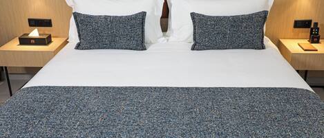 Quarto de hotel com uma cama grande, dois travesseiros brancos, dois travesseiros azuis texturizados, um cobertor azul e duas mesas de cabeceira com luminárias e uma caixa de lenços.