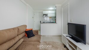 Uma sala de estar com um sofá bege, uma televisão de tela plana e uma cozinha ao fundo.