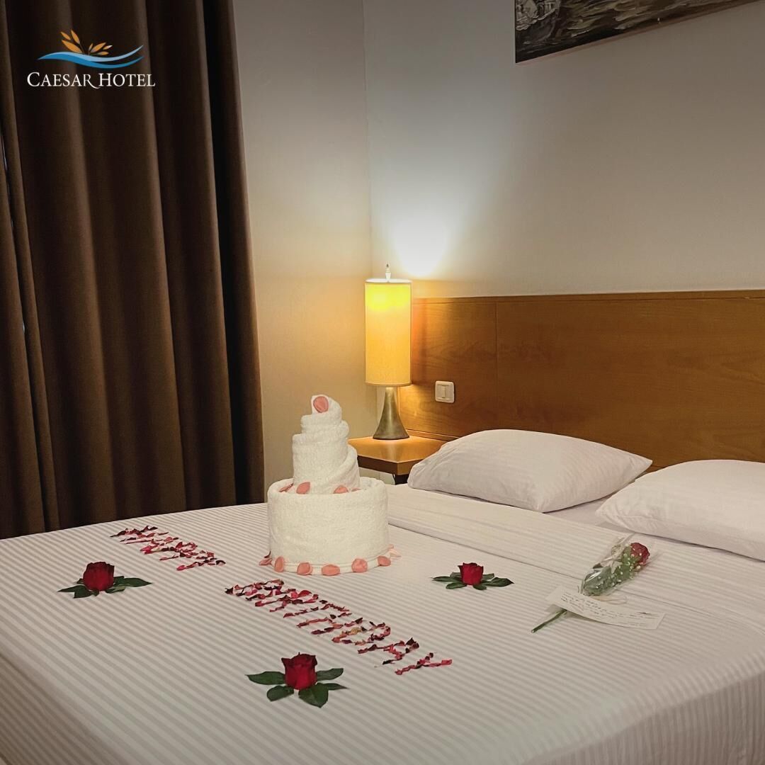Quarto de hotel com uma cama, mesa de cabeceira, luminária e um bolo com a mensagem 'Feliz Aniversário'.