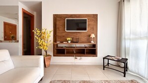 Uma sala de estar com um sofá branco, um móvel de televisão em madeira, uma mesinha e um vaso com planta.