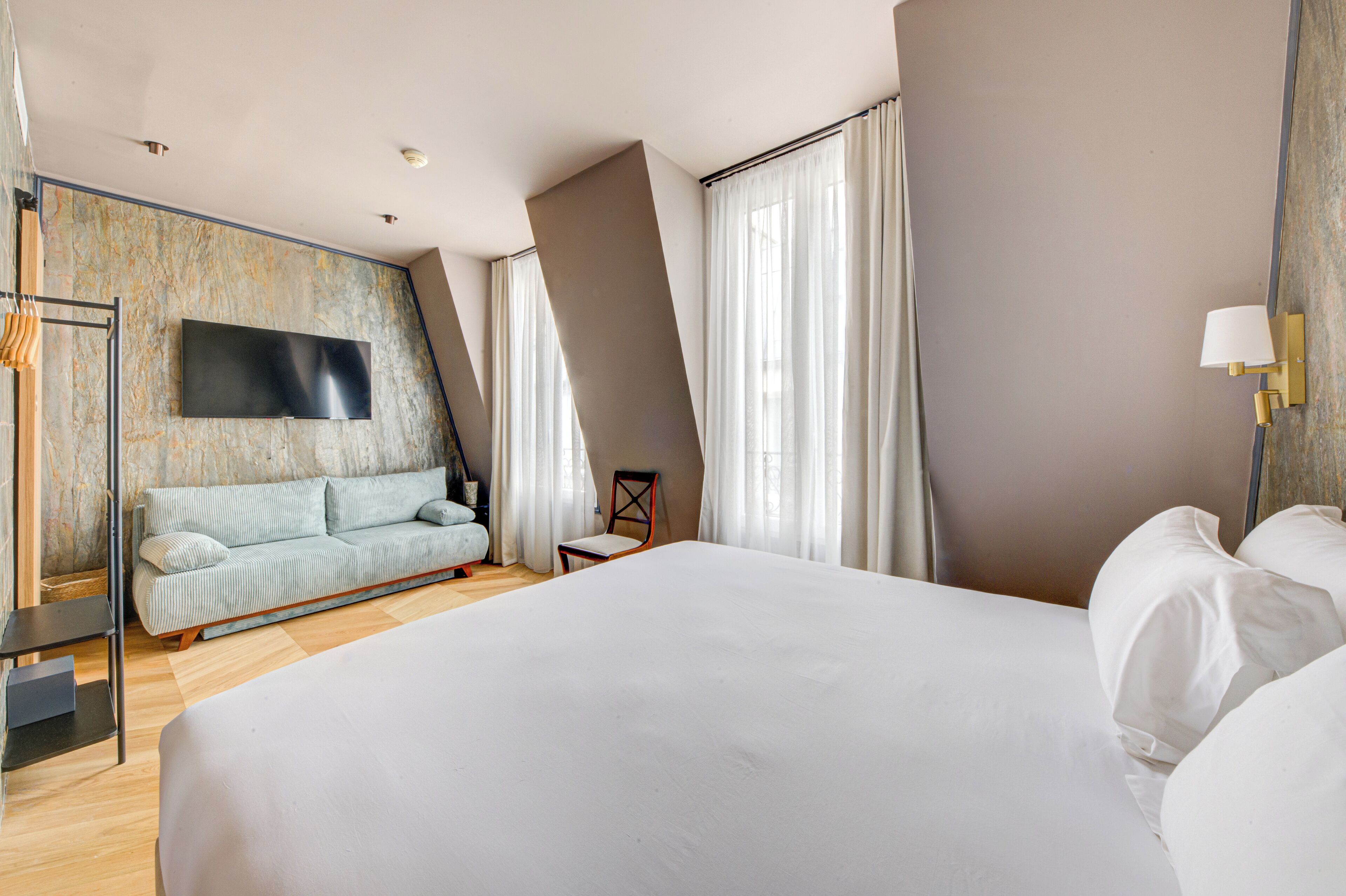 Quarto de hotel moderno com uma cama grande, um sofá, uma TV de tela plana na parede texturizada e uma janela com cortinas translúcidas.