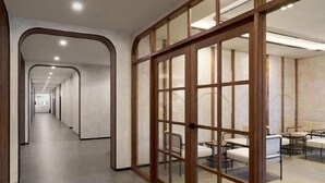 Corredor moderno de hotel com divisória de madeira, que leva a uma área de estar com mesas e cadeiras.