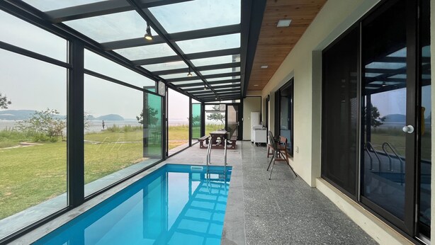 Piscina interior moderna com grandes janelas, teto de madeira e vista para área ajardinada e corpo d'água.