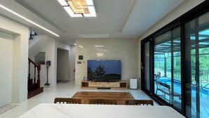 Uma sala de estar moderna com uma grande televisão de tela plana em um suporte de madeira, uma área de jantar com uma mesa de mármore e uma grande porta de vidro deslizante que dá para uma área externa.