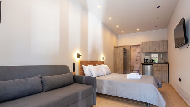 Quarto de hotel moderno com sofá cinzento, cama com cabeceira de madeira, pequeno kitchenette e televisão fixada na parede.