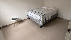 Um quarto pequeno com uma cama de solteiro, uma colcha branca, uma pequena mesa de cabeceira e um ar condicionado fixado na parede.