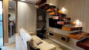 Uma sala de estar moderna com um sofá, uma TV fixada em uma prateleira de madeira e uma porta deslizante para uma varanda.